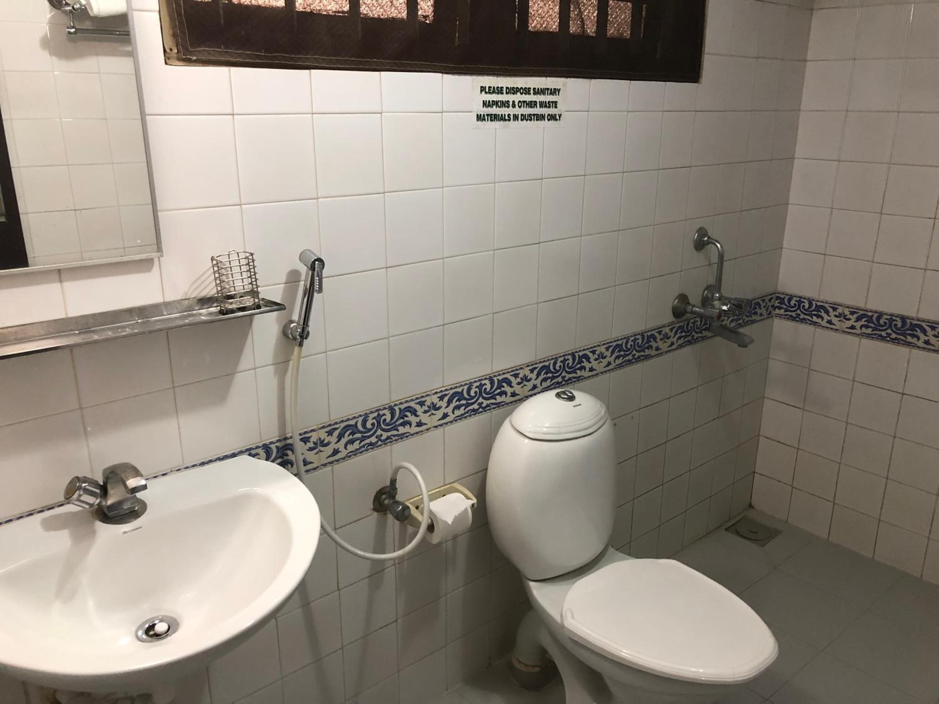 Uma casa de banho com lavatório, sanita e chuveiro. Um aviso indica para deitar resíduos sanitários num contentor de lixo.
