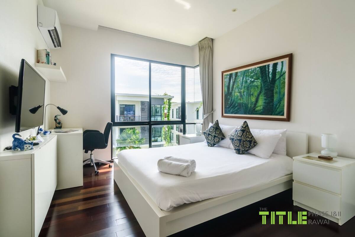 Quarto de hotel moderno com uma cama grande, uma escrivaninha, uma cadeira, televisão e janela com vista para edifícios e vegetação.