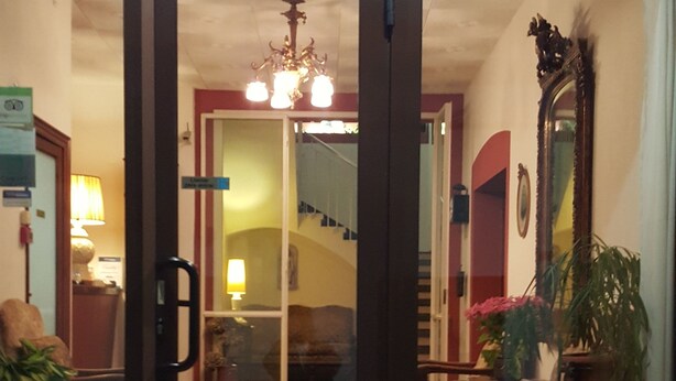 Entrada de hotel com porta de vidro, lustre, planta em vaso e escadaria para o interior.