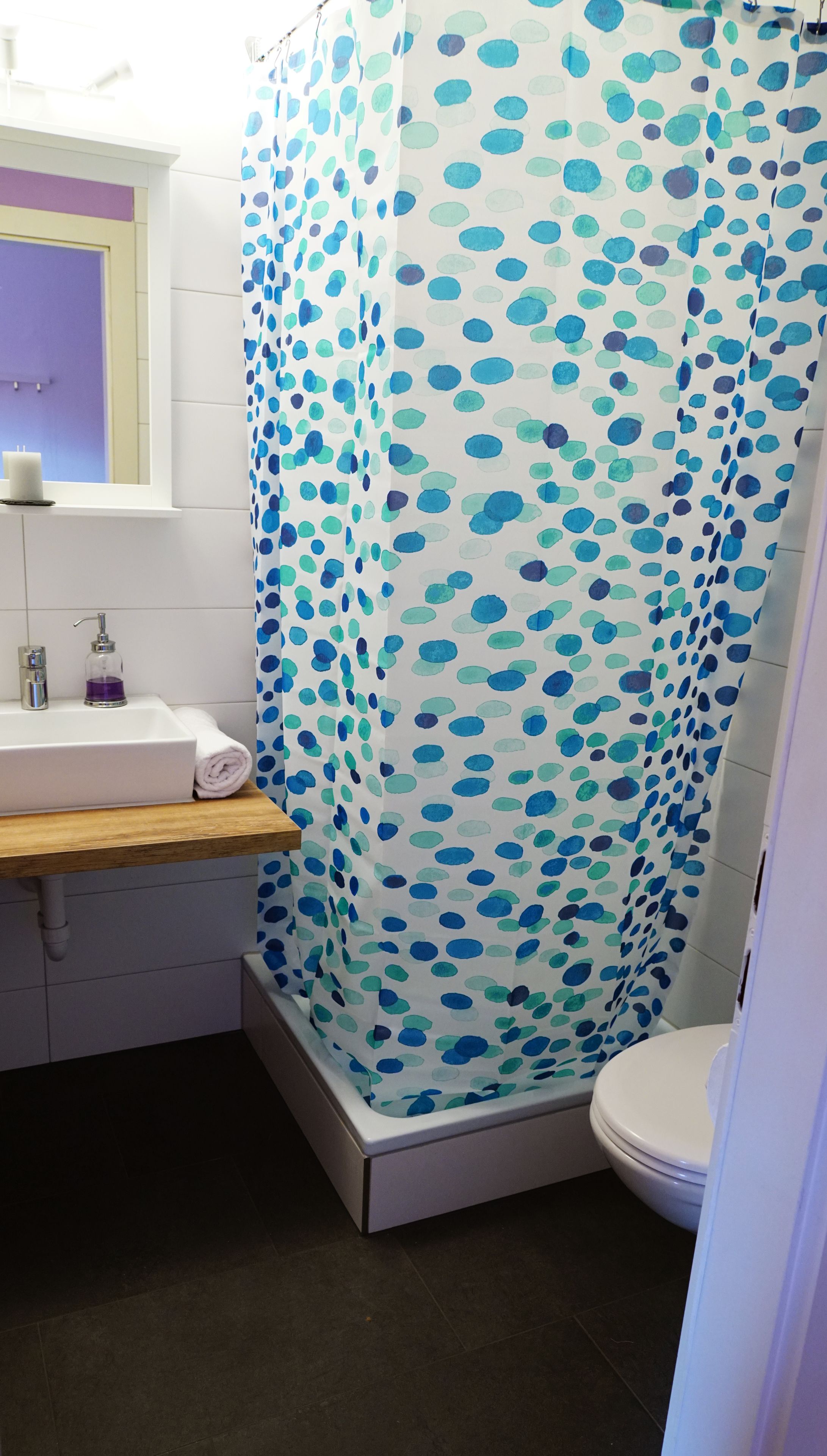 Um banheiro com cortina de chuveiro estampada em bolinhas azuis e brancas, pia branca com dispensador de sabonete e vaso sanitário.