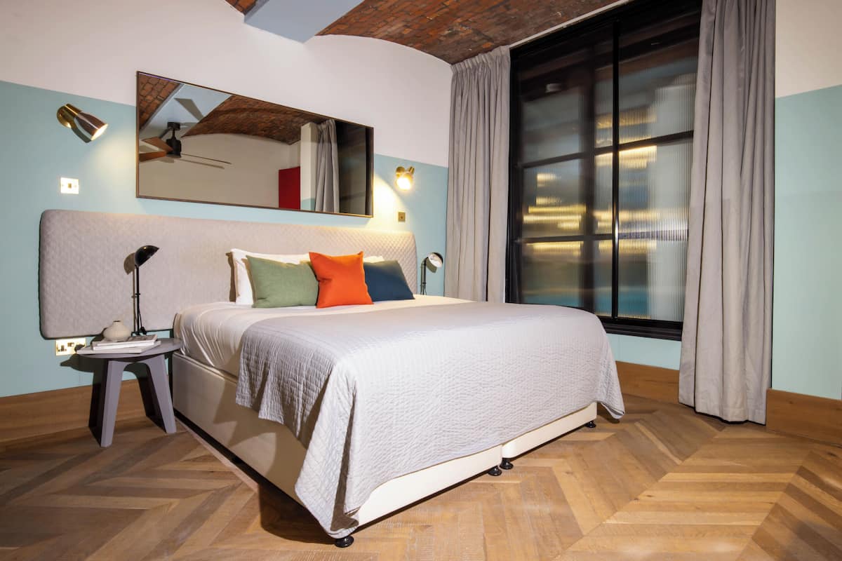 Quarto com cama, mesa de cabeceira, espelho e uma janela grande com cortinas.