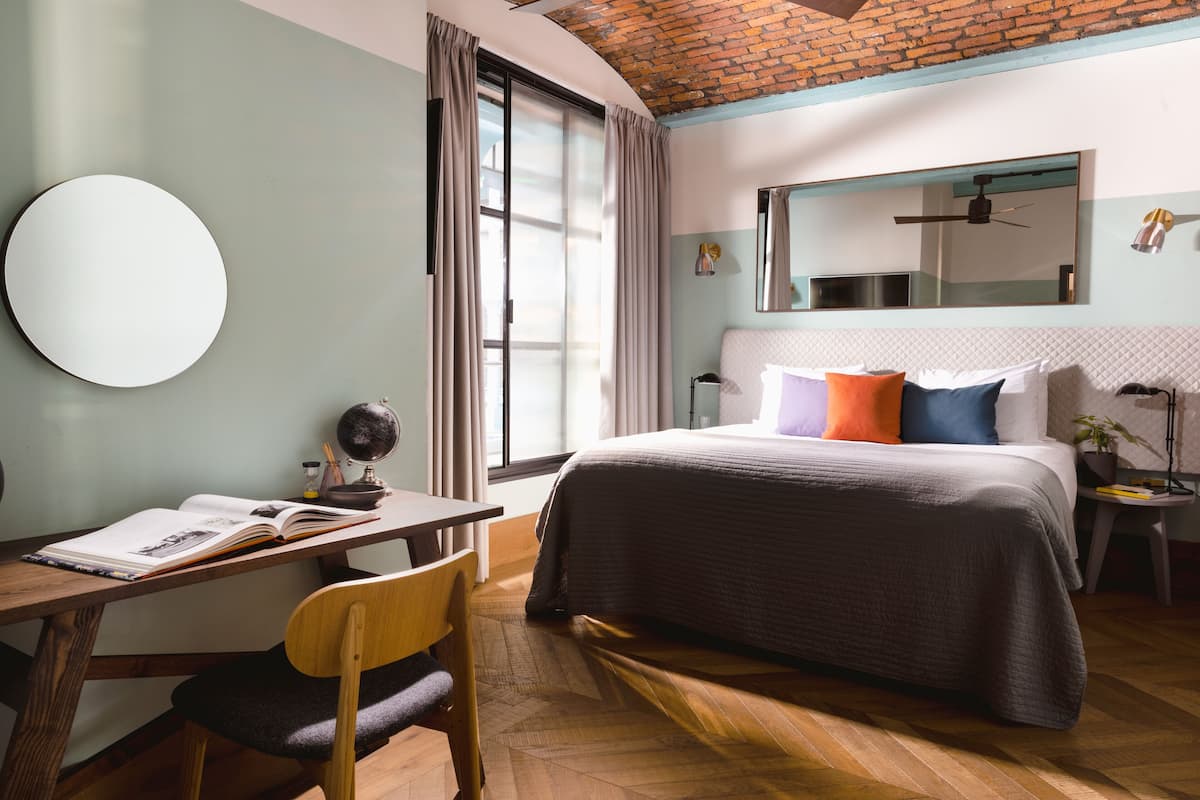 Quarto de hotel com uma cama grande, uma escrivaninha com cadeira, um espelho redondo e uma janela com cortinas.