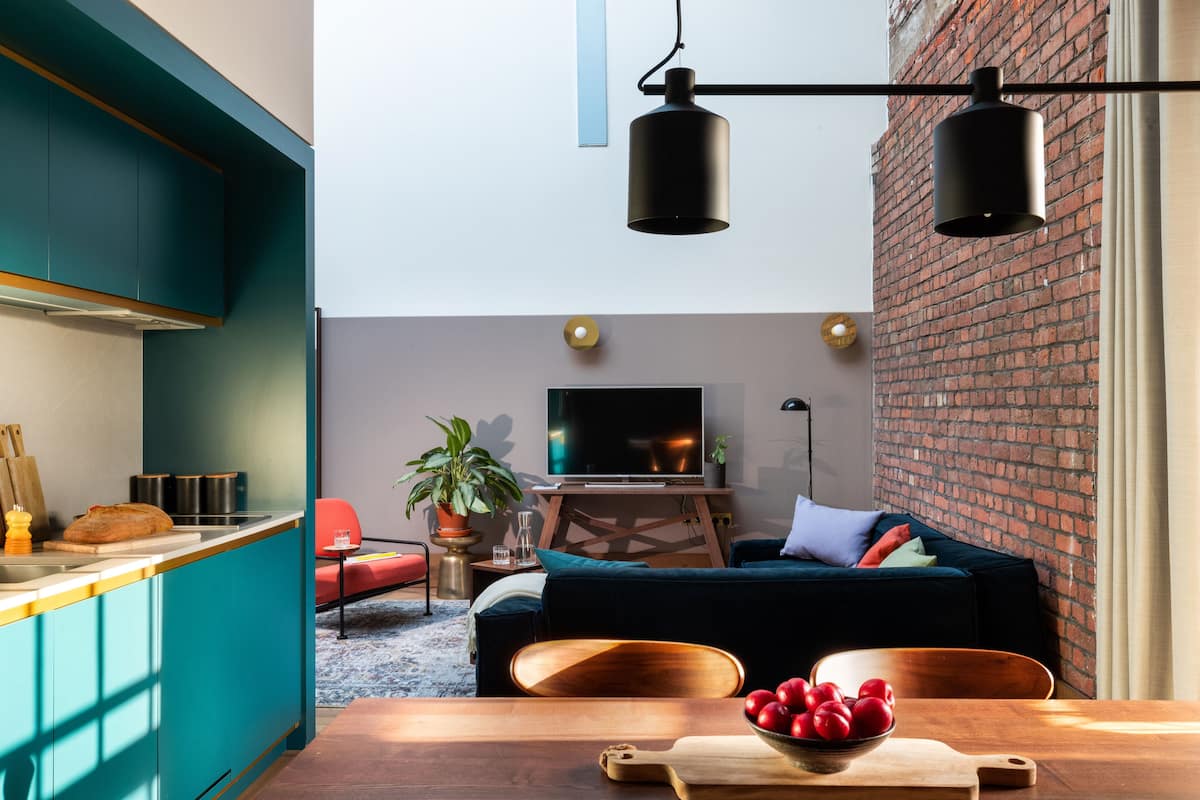Uma cozinha moderna com armários verde-água, uma sala de jantar com mesa de madeira e uma sala de estar com sofá azul-escuro e uma TV.