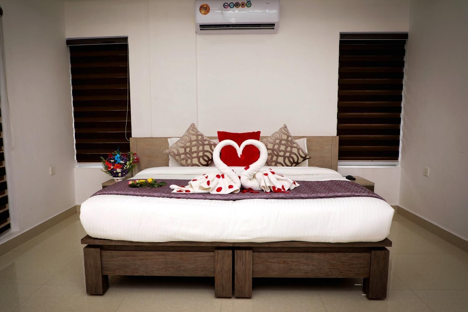 Um quarto com uma cama grande, duas janelas com persianas, um aparelho de ar condicionado e um buquê de flores na mesa de cabeceira.