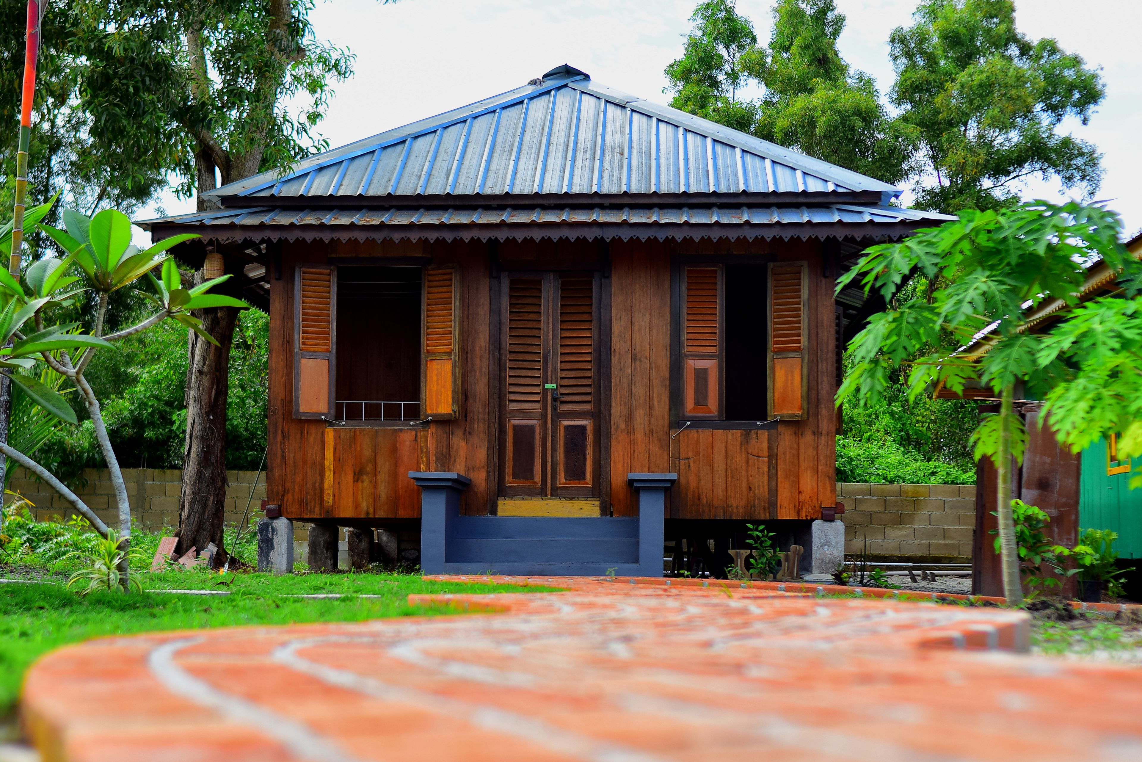 Uma casa de madeira tradicional com telhado de metal, um caminho de tijolos até a entrada e vegetação exuberante ao redor.