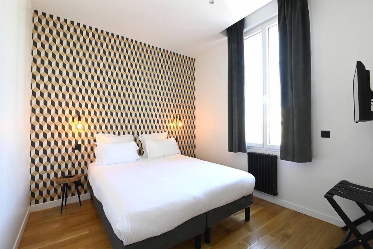 Quarto com papel de parede estampado, cama com roupa de cama branca, um pequeno banquinho de madeira, radiador e janela com cortinas.
