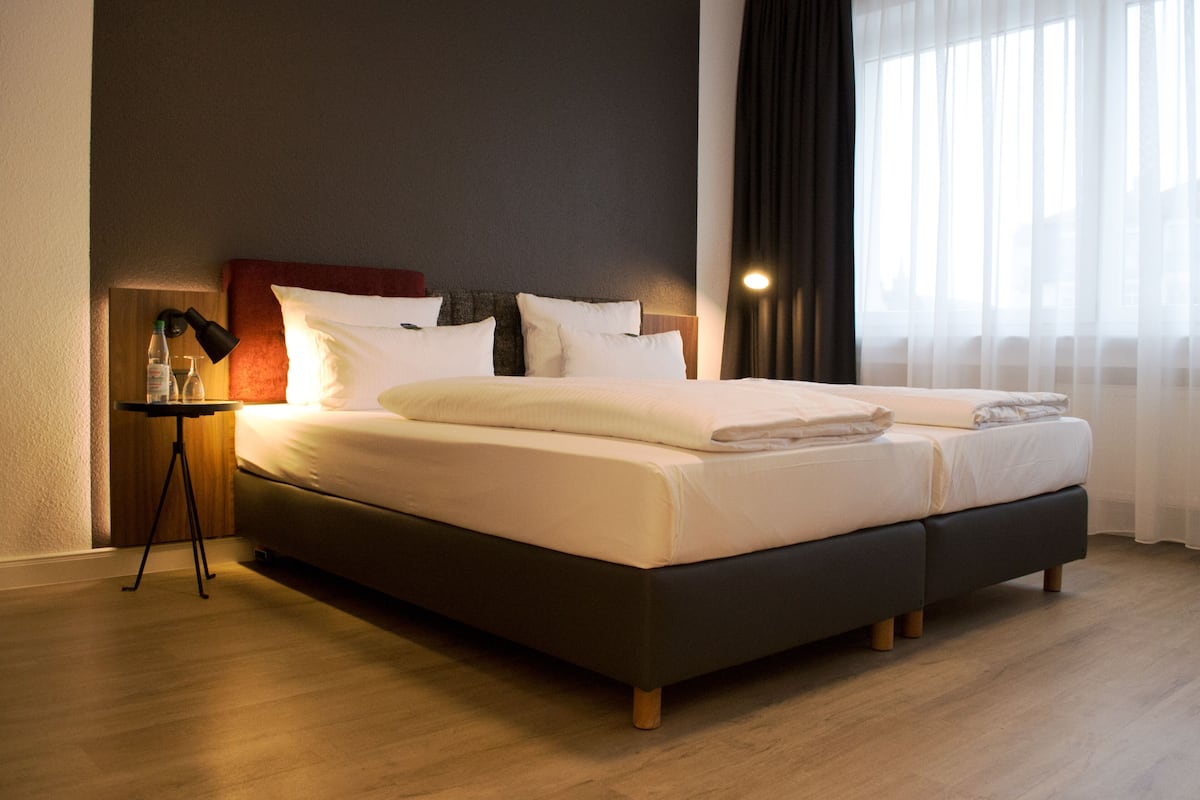 Uma cama bem arrumada com roupa de cama branca, uma mesa de cabeceira com abajur e uma janela com cortinas finas.