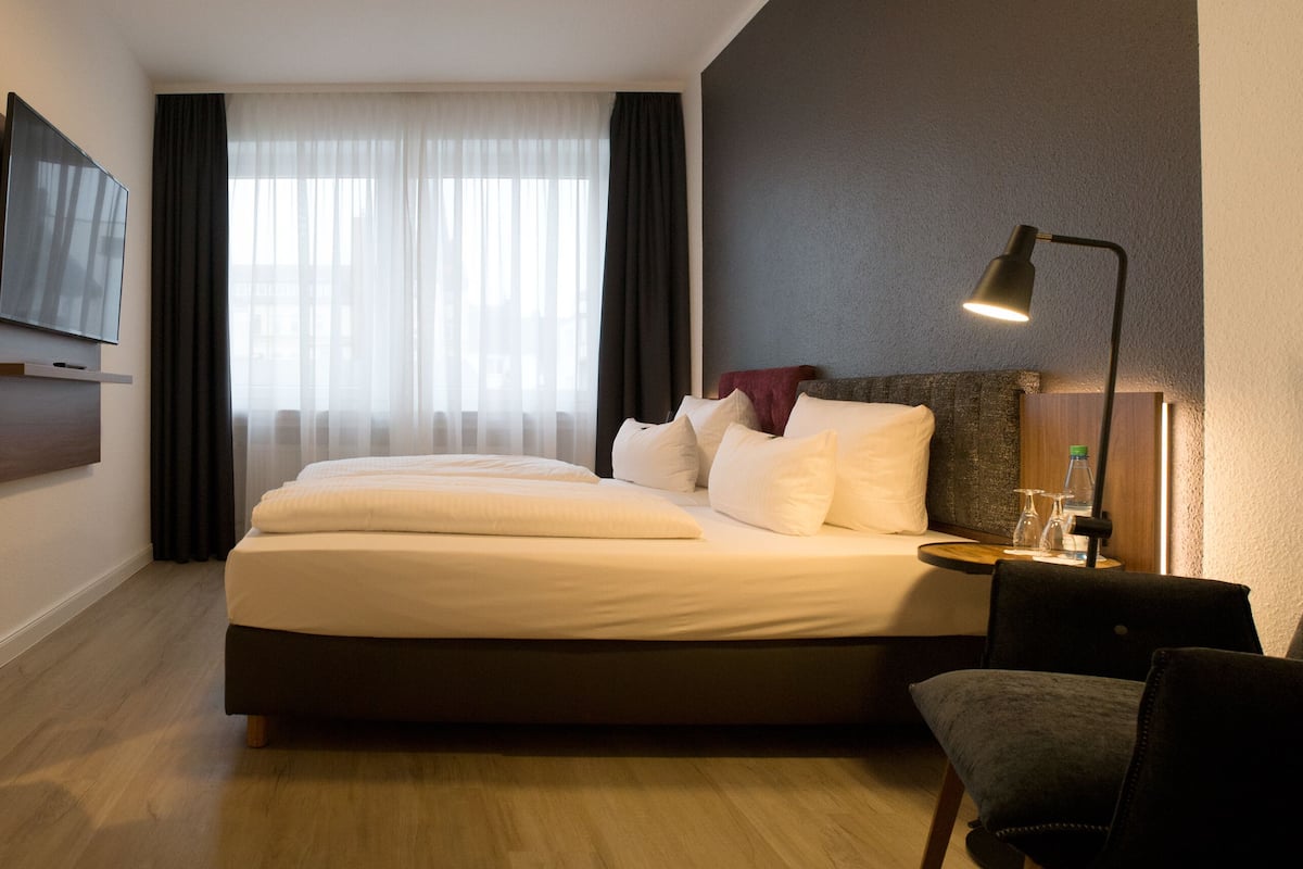 Quarto de hotel com uma cama, televisão na parede, mesa de cabeceira com copos e uma luminária.