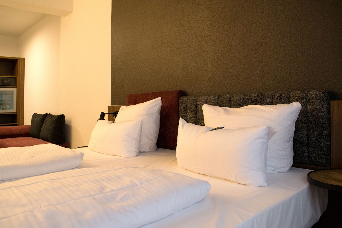 Quarto de hotel com duas camas, roupa de cama branca, cabeceira marrom e uma pequena mesa de cabeceira de madeira.
