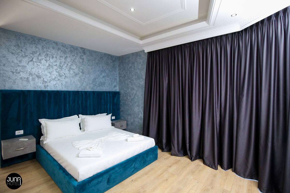 Quarto de hotel moderno com uma cama grande, uma mesa de cabeceira e uma cortina escura.