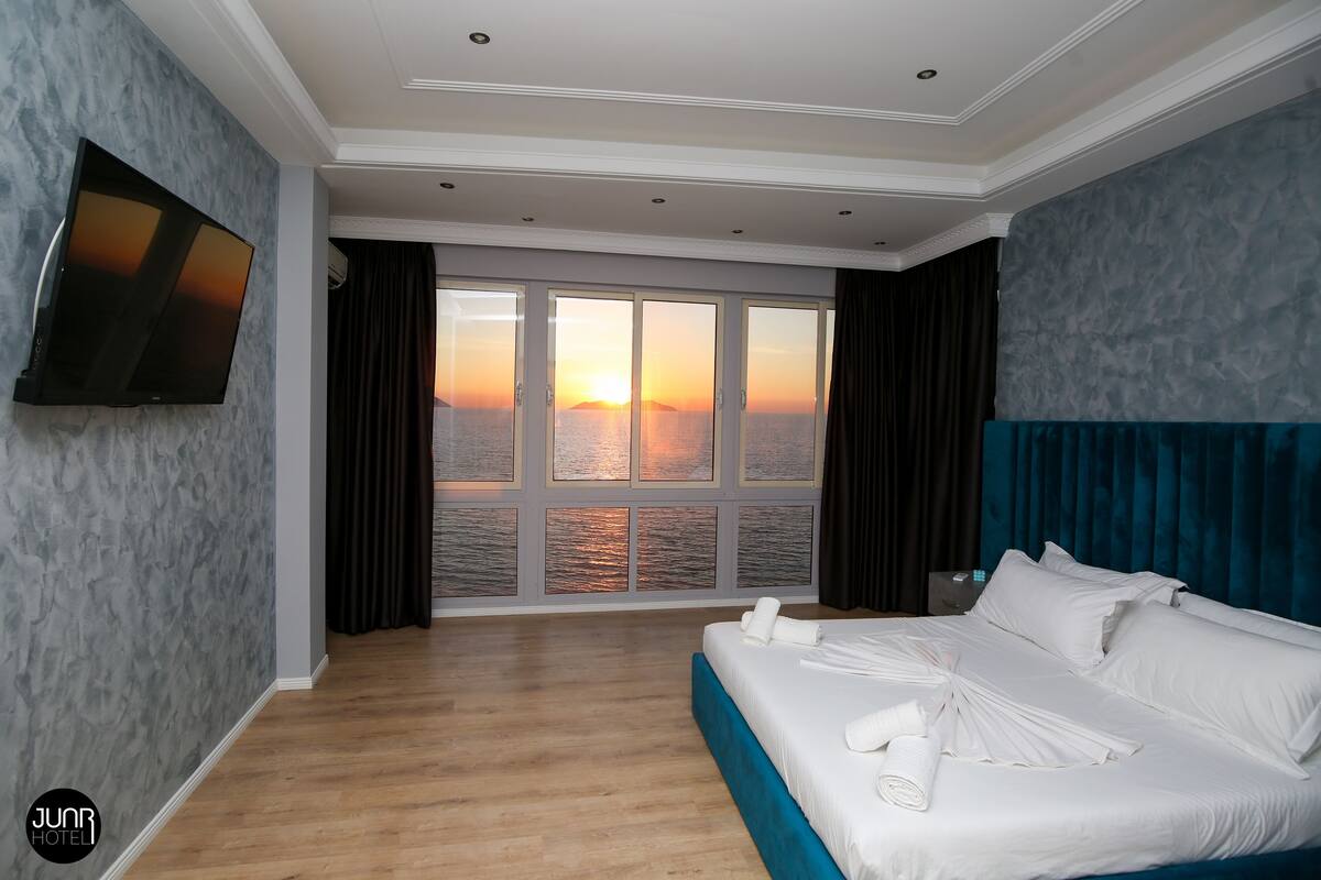 Quarto de hotel com uma cama grande, televisão de tela plana e vista do pôr do sol sobre a água.