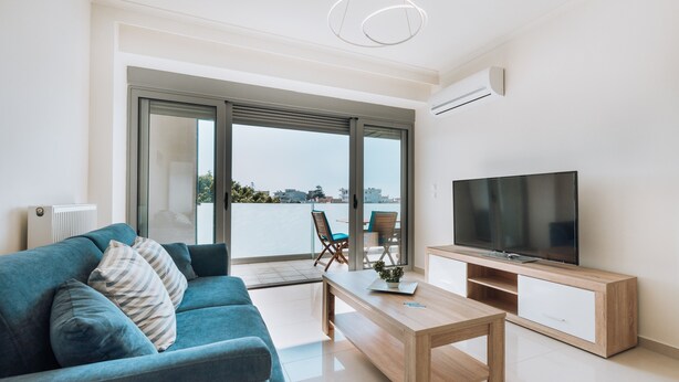 Uma sala de estar moderna com um sofá azul, uma mesa de centro, uma TV sobre um suporte de madeira e uma varanda com vista para edifícios.