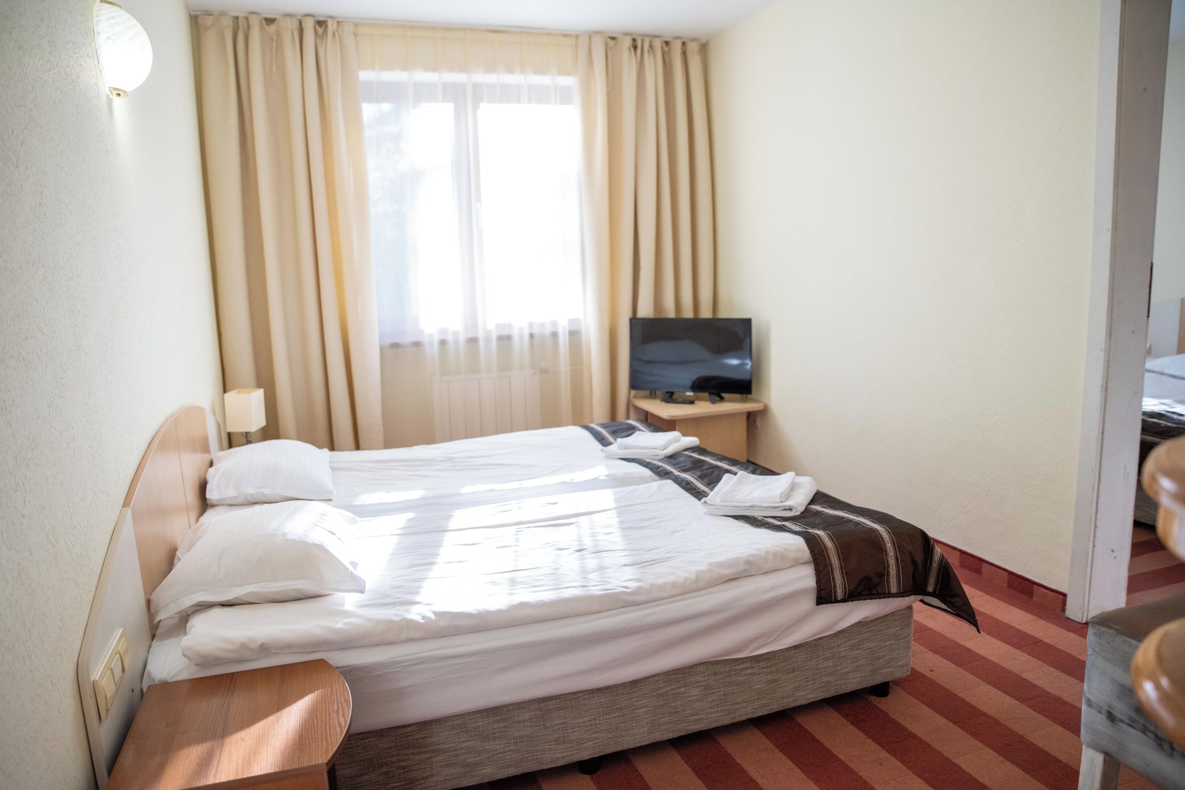 Quarto de hotel com cama, televisão, mesa de cabeceira e janela com cortinas.