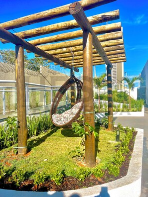 Uma cadeira suspensa sob uma pérgula de bambu em um jardim bem cuidado, com vista para um edifício e um céu limpo.