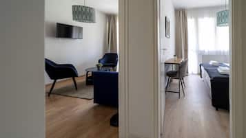 Quarto moderno e bem iluminado com piso de madeira, televisão de tela plana na parede, uma mesinha redonda com uma planta, uma cadeira azul, uma cama com cabeceira preta e uma janela com cortinas leves.