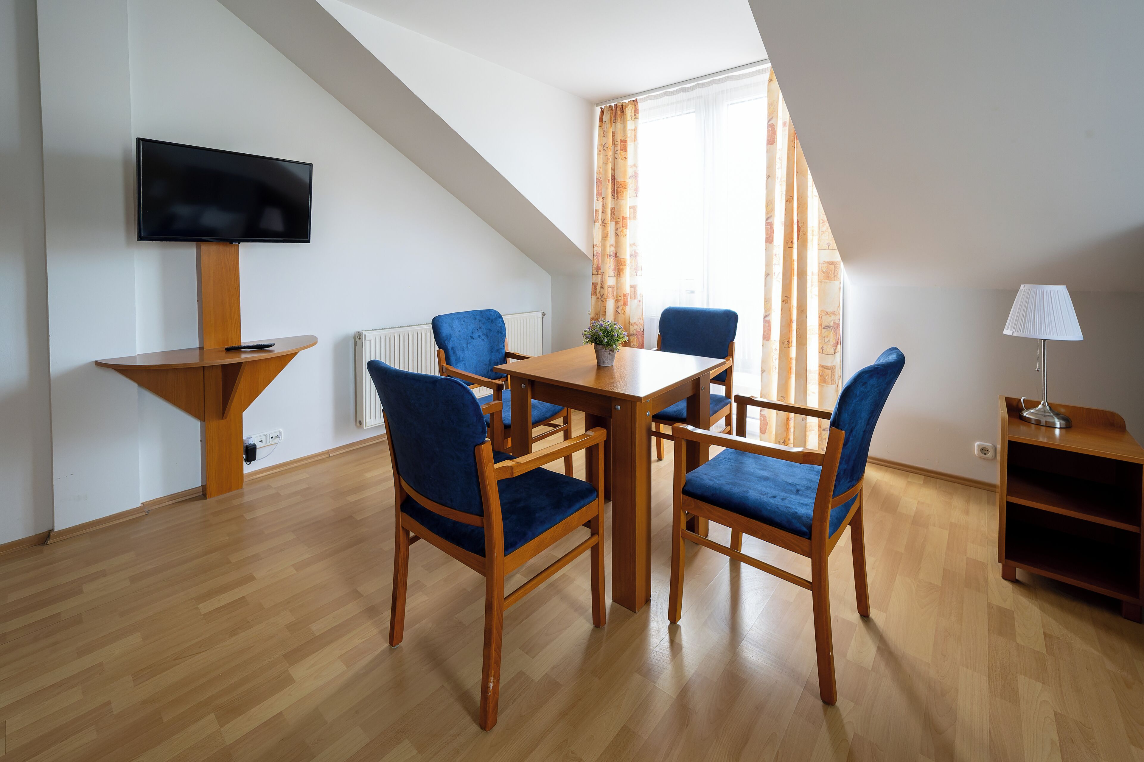 Uma sala de estar moderna com uma mesa de madeira, cadeiras azuis, uma televisão de tela plana, uma luminária e uma janela com cortinas.
