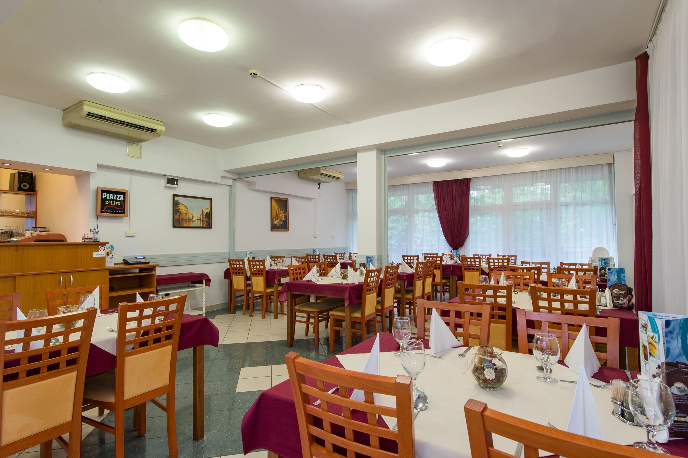 Uma área de refeições com várias mesas e cadeiras, uma cozinha com uma placa que diz “PIAZZA D’Oro”, e uma grande janela com cortinas.