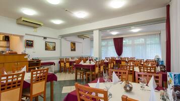 Uma área de refeições com várias mesas e cadeiras, uma cozinha com uma placa que diz “PIAZZA D’Oro”, e uma grande janela com cortinas.