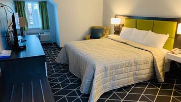 Quarto de hotel com cama, mesa de cabeceira, luminária e escrivaninha com computador e telefone.