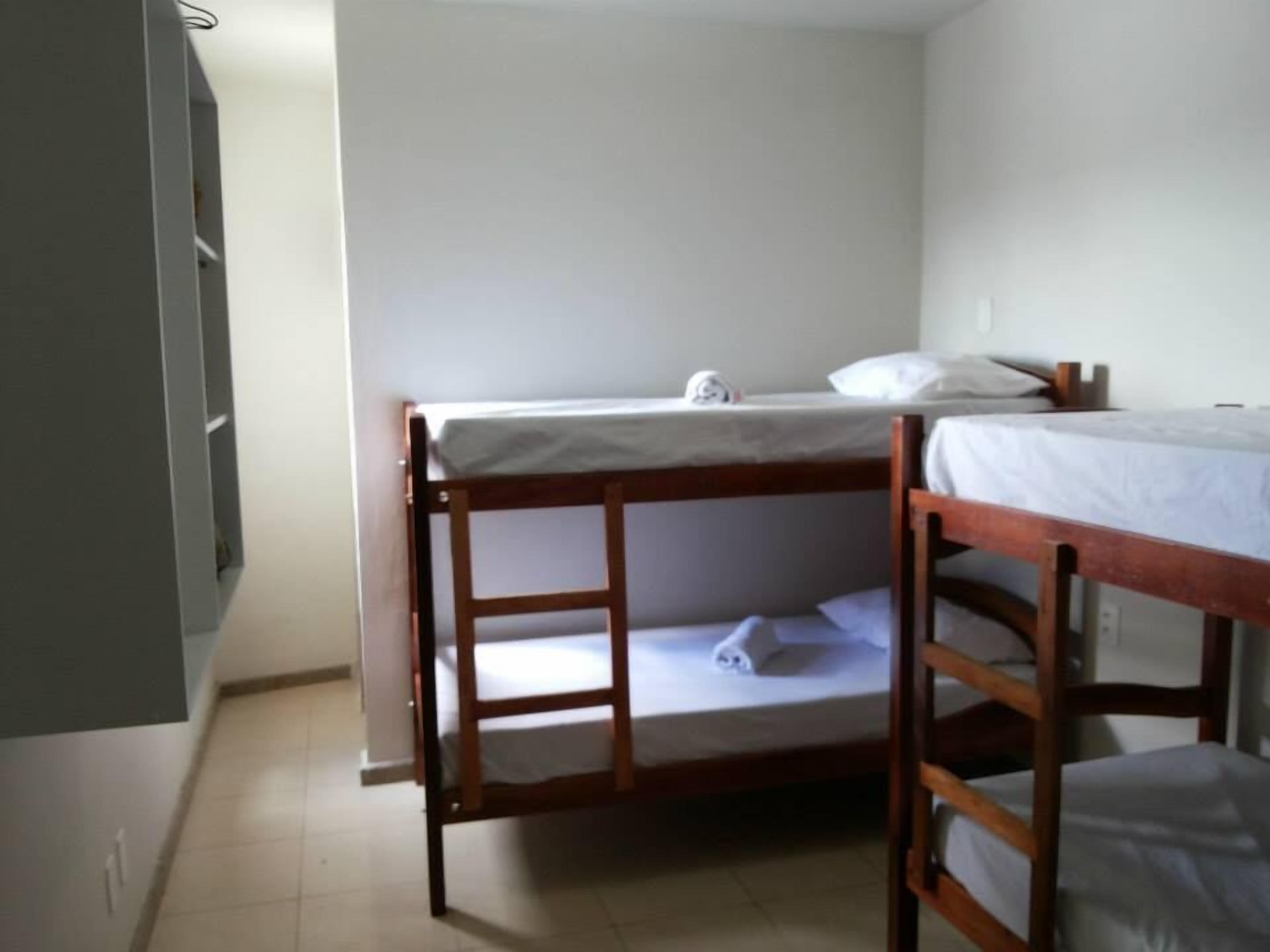 Quarto pequeno e compacto com uma cama de beliche e uma cama simples. Um guarda-roupa de madeira está localizado no lado esquerdo.