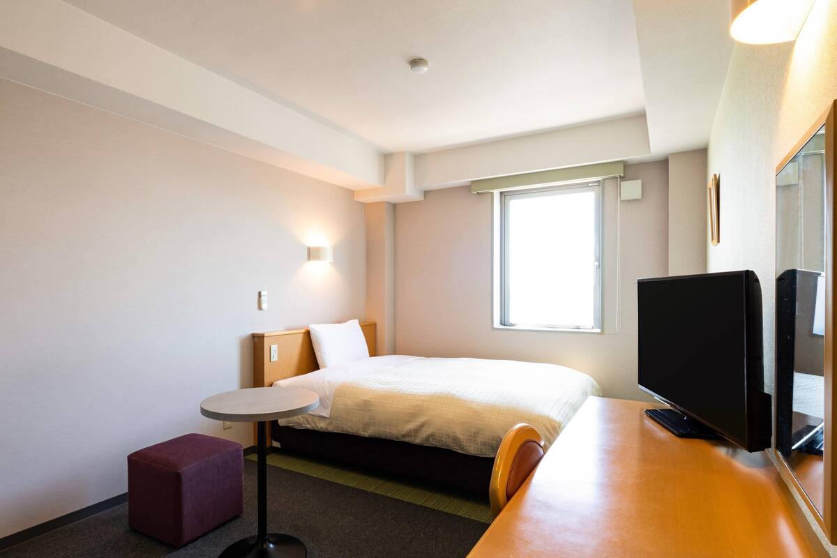 Quarto de hotel com uma cama, uma mesa pequena, uma televisão e uma janela.