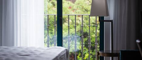 Quarto com cama, mesa de cabeceira, luminária, cadeira e varanda com vista para o verde.