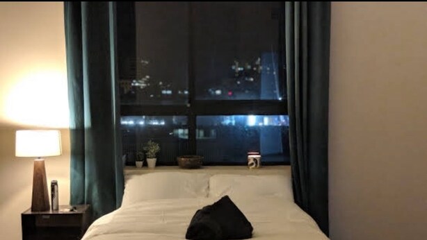 Quarto de hotel com cama, mesa de cabeceira, luminária, janela com cortinas e vista da cidade à noite.