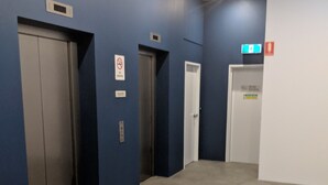 Corredor com paredes azuis, chão de azulejos e três portas, uma delas saída de emergência.