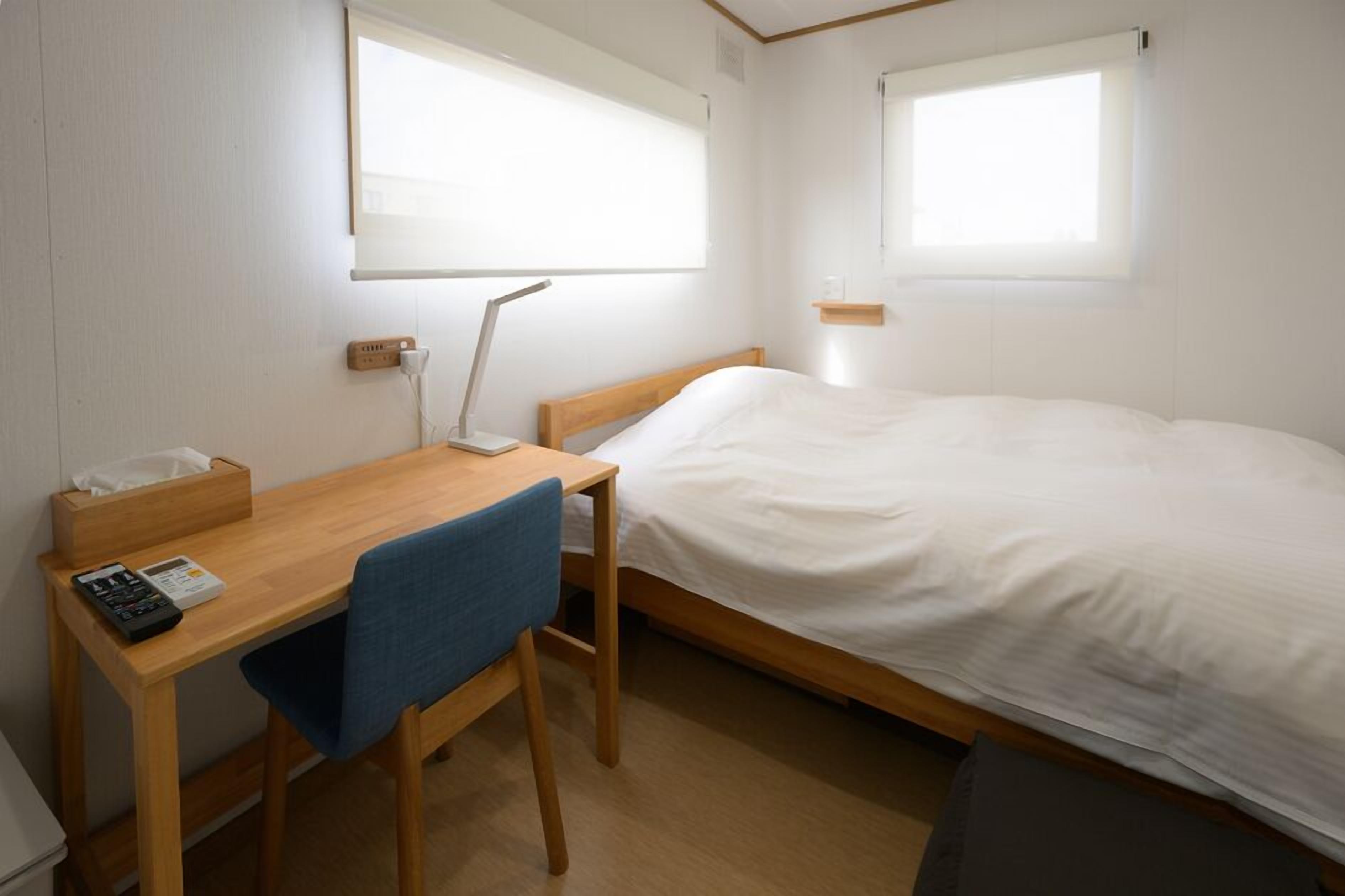 Quarto pequeno com uma cama de solteiro, escrivaninha de madeira, cadeira, mesa de cabeceira e janela.