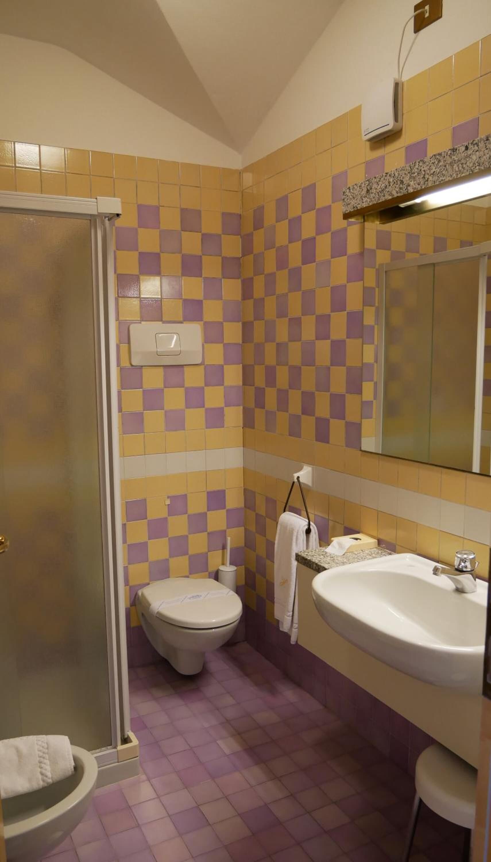 Um banheiro com parede de azulejos quadriculada em amarelo e roxo, vaso sanitário branco, pia e espelho.