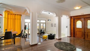 Ampla sala de estar com mesa de jantar, cadeiras, sofá e mesa de centro. Há uma porta de madeira com painéis de vidro e uma luminária pendente moderna.