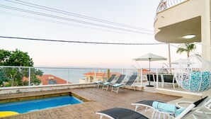 Uma área de piscina no terraço com espreguiçadeiras, guarda-sóis e vista para o mar.