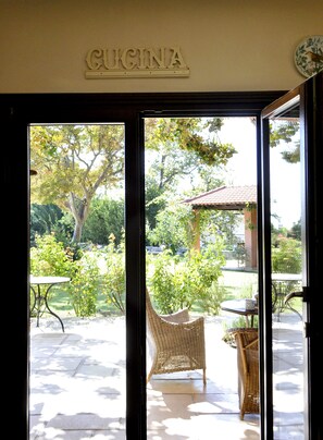 Uma entrada com uma placa indicando "CUGINA" leva a um pátio ao ar livre com móveis de vime e vista para um jardim e um edifício com telhado de azulejos.