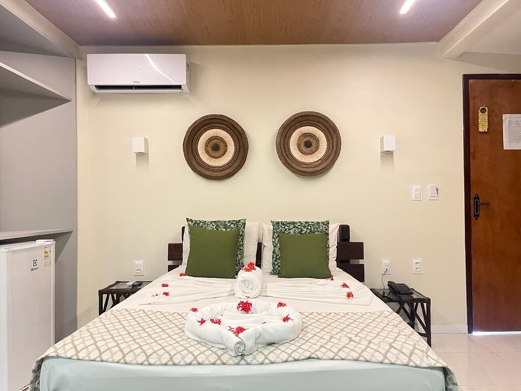 Quarto de hotel com uma cama, dois aparelhos de ar condicionado fixados na parede, uma porta de madeira e uma pequena mesa com um telefone.