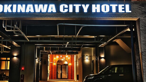 Exterior do Hotel Okinawa City, com uma placa bem visível, entrada moderna e um carro estacionado.