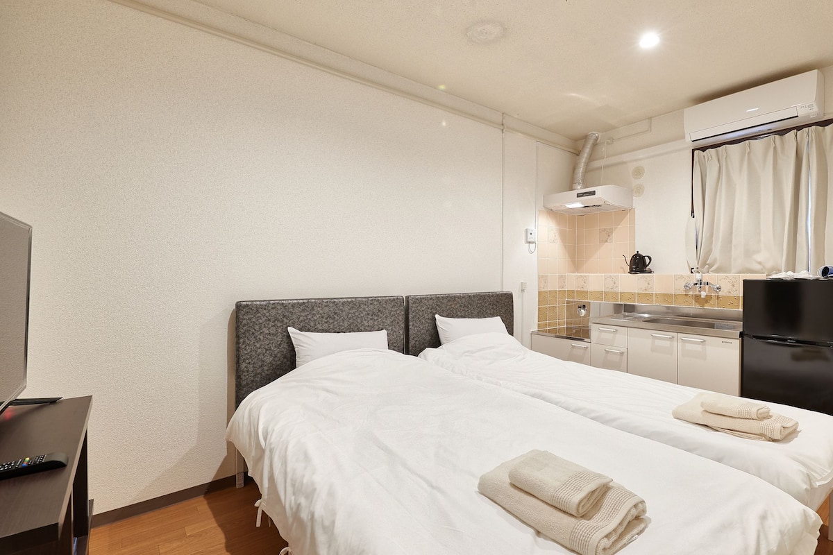 Quarto de hotel com duas camas, televisão, secretária, cozinha compacta e casa de banho.
