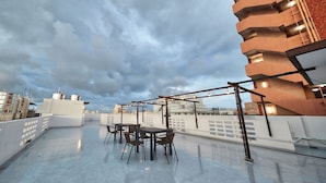 Terraço no telhado com mesa e cadeiras, pérgola com luzes de corda e vista dos edifícios ao redor.