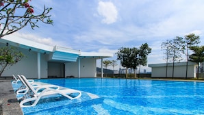 Piscina com cadeiras de descanso brancas, céu azul límpido e um edifício moderno com telhado plano ao fundo.