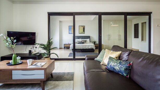 Sala de estar moderna com sofá, mesa de centro e televisão. Um quarto é visível através de uma porta de vidro.