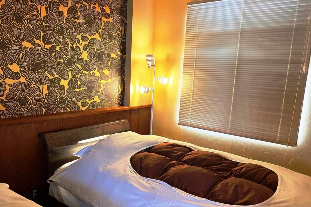 Uma cama de solteiro com cabeceira, uma luminária fixada na parede e uma janela com persianas.