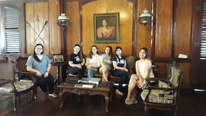 Um grupo de pessoas sentadas em um sofá e cadeiras de madeira em um ambiente com painéis de madeira e uma grande moldura na parede.