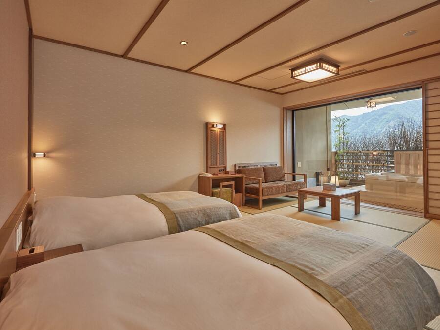 Quarto japonês tradicional com tatames, duas camas, uma mesa baixa e uma porta deslizante que dá para uma varanda com vista para a montanha.
