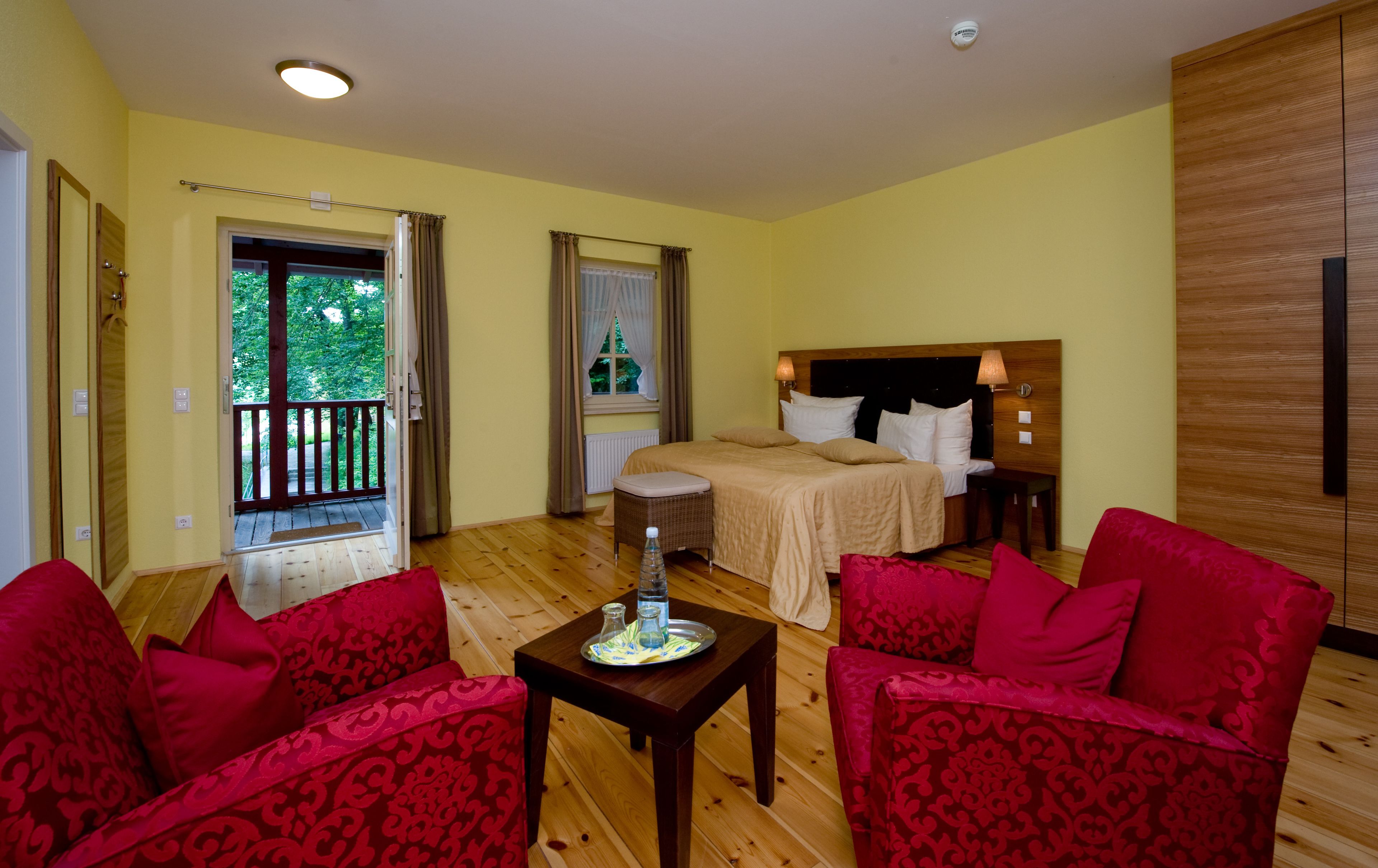 Quarto de hotel com uma cama, duas cadeiras vermelhas estampadas, uma mesa pequena com uma garrafa e copos, um guarda-roupa de madeira e uma varanda com guarda-corpo.