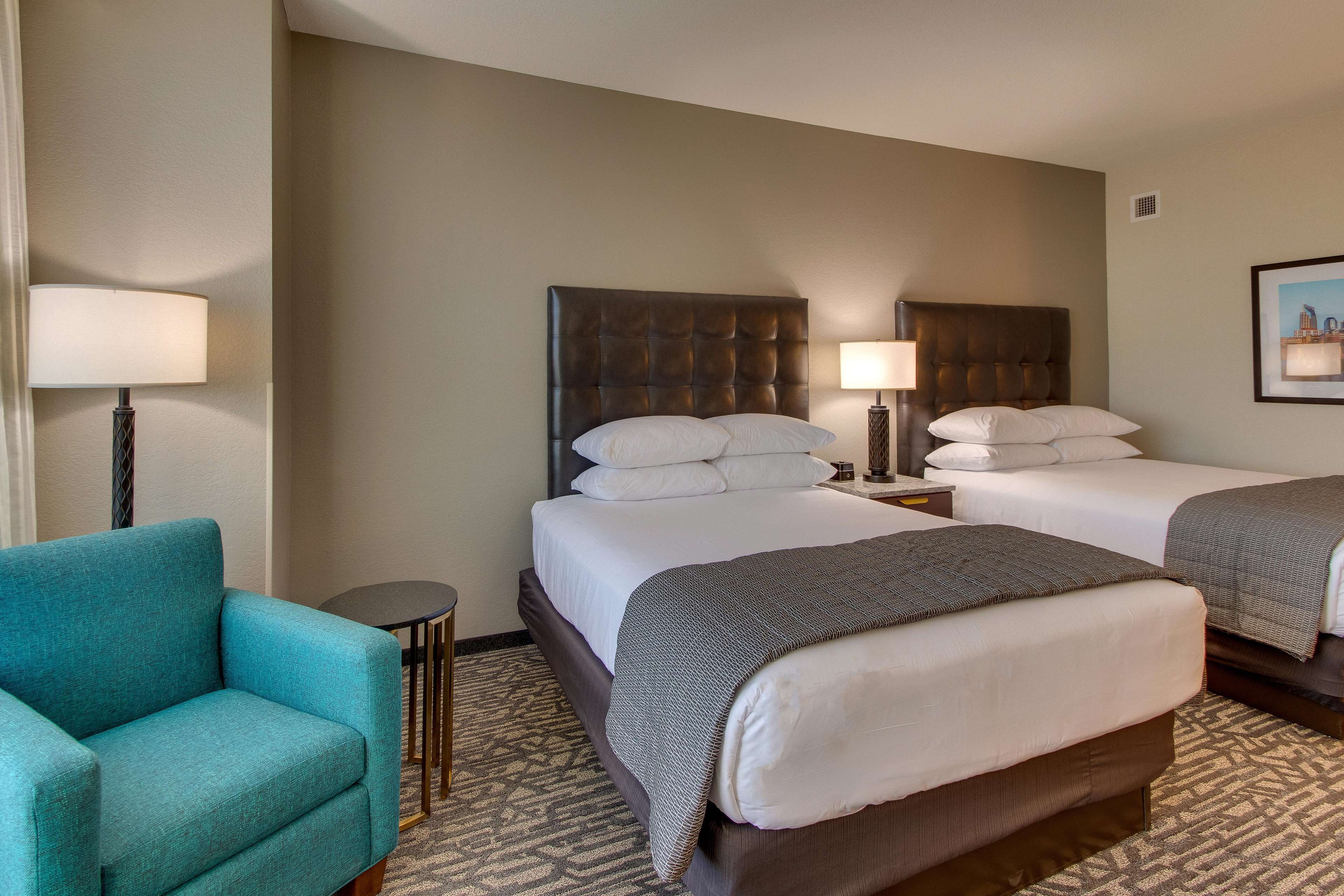 Quarto de hotel com duas camas, uma poltrona azul, uma mesinha e uma luminária.