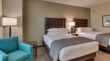 Quarto de hotel com duas camas, uma poltrona azul, uma mesinha e uma luminária.