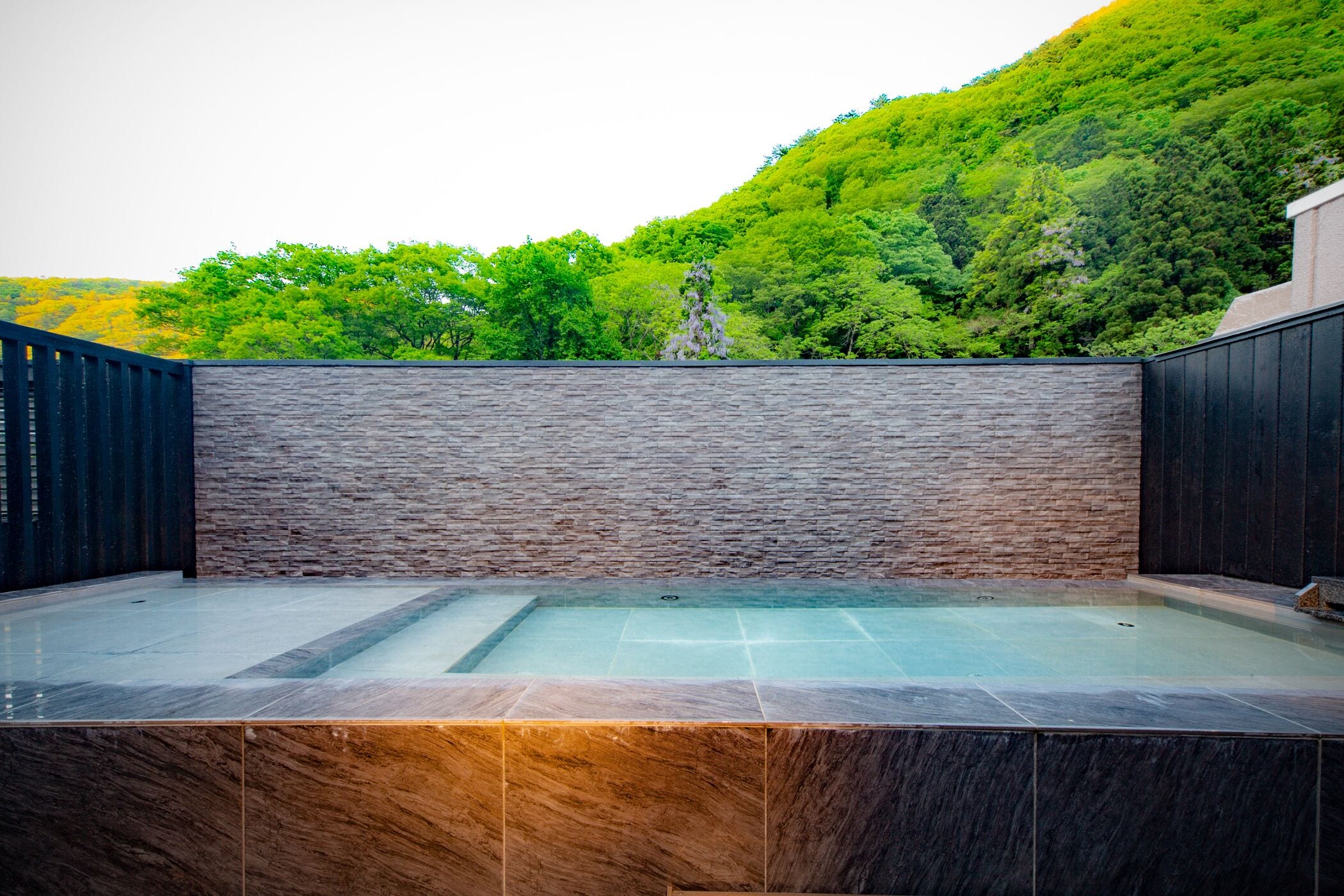 Uma área de spa com parede de pedra, piscina e vista para uma encosta verde exuberante.