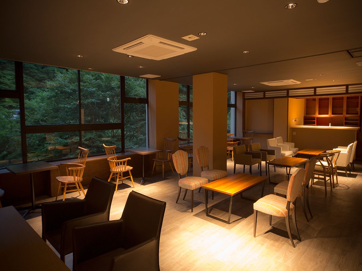 Uma sala de jantar moderna com mesas e cadeiras de madeira, amplas janelas com vista para árvores e iluminação embutida no teto.