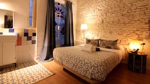 Quarto com cama grande, parede de pedra, janela com cortinas e mesa de cabeceira com abajur.