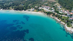 Vista aérea de uma vila costeira com casas ao longo da orla, mar azul cristalino e montanhas ao fundo.