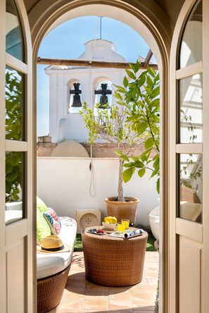 Uma área de estar externa acolhedora com mesa de vime, uma árvore em vaso e vista para um edifício branco com uma torre de sino.